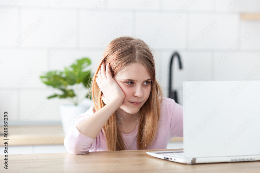 Puzzled teen girl sitting at the table using laptop studying online at ...
