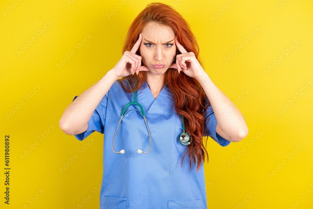 Serious concentrated young red-haired doctor woman over yellow studio ...