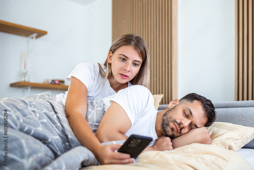 Jealous wife spying the phone of her partner while he is sleeping in a bed at home. Shocked ...