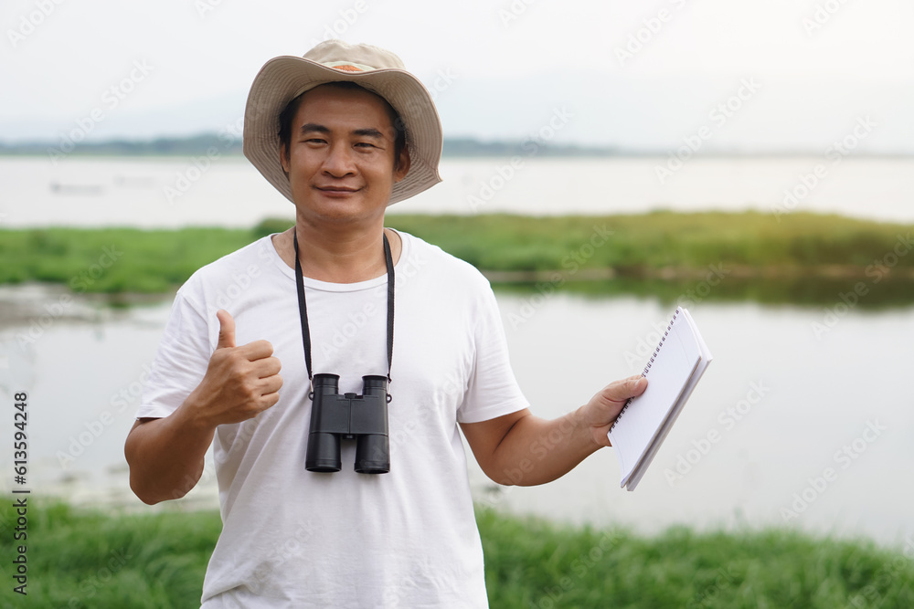 Handsome Asian man ecologist is surveying nature at the lake, holds paper notebook and ...