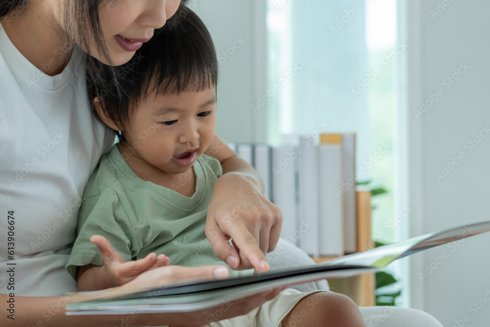 Happy Asian mother relax and read book with baby time together at home. parent sit on sofa with ...
