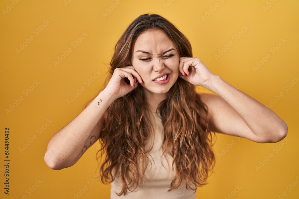 Young hispanic woman standing over yellow background covering ears with ...