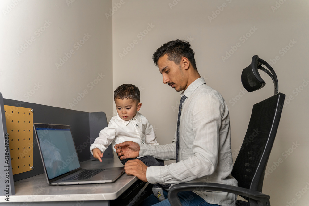 Father taking care of his son while working at a desk. Working parent ...