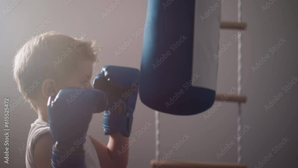 Concentrated little boy in gloves punches bag hanging in gym by rope ...