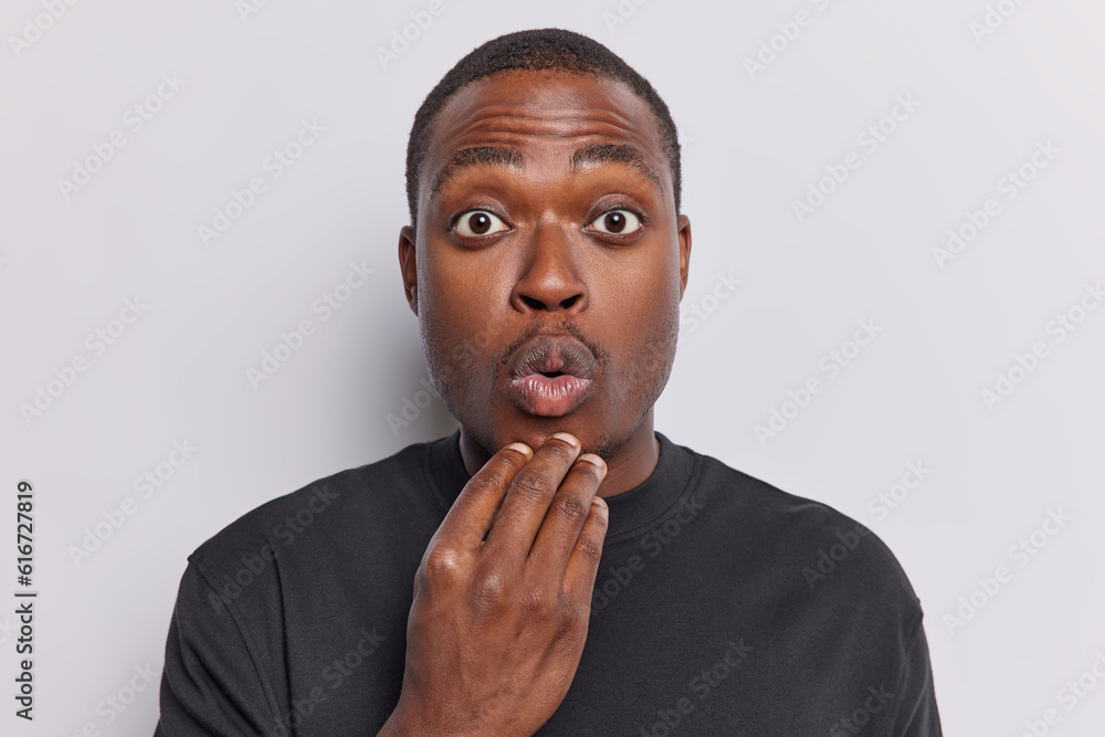 Portrait of dark skinned man keeps hand on chin lips rounded reacts to ...