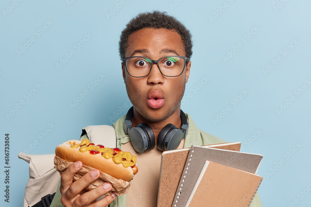 Shocked male student poses with notebooks and hot dog has snack during ...