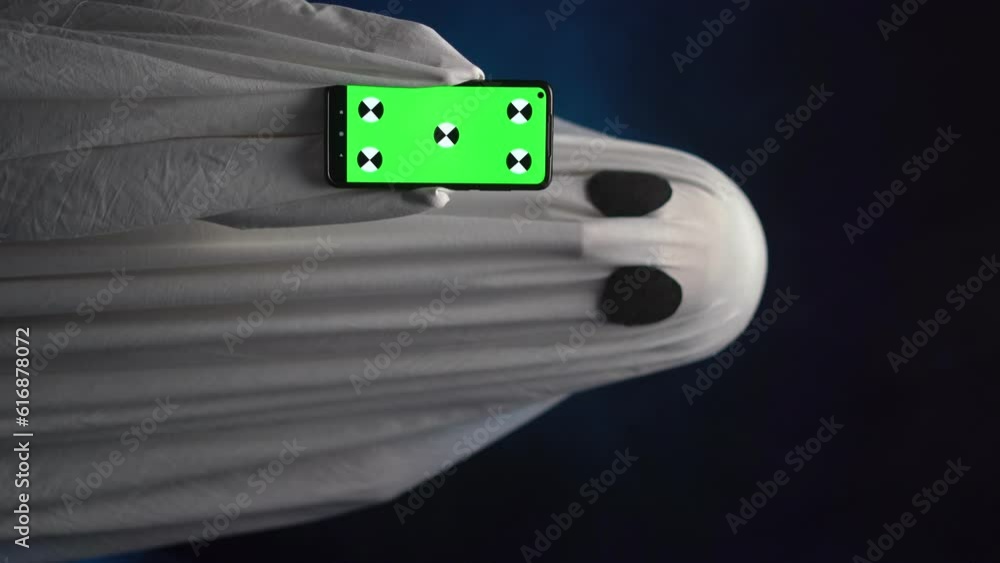 Ghost in white sheet holding hand up shows mobile smartphone with green ...