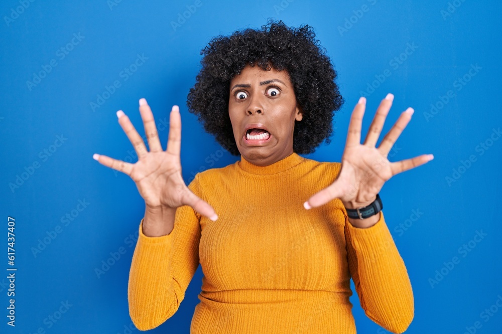 Black woman with curly hair standing over blue background afraid and ...