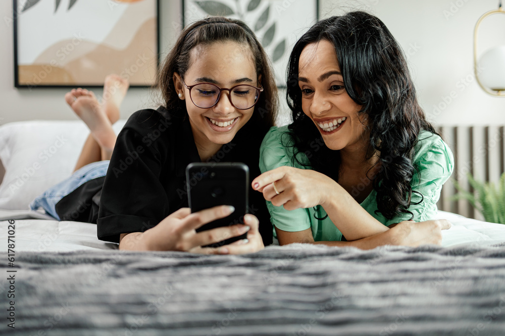 Mother and teenage daughter using smartphone together lying in bed at home. Latina mother and ...