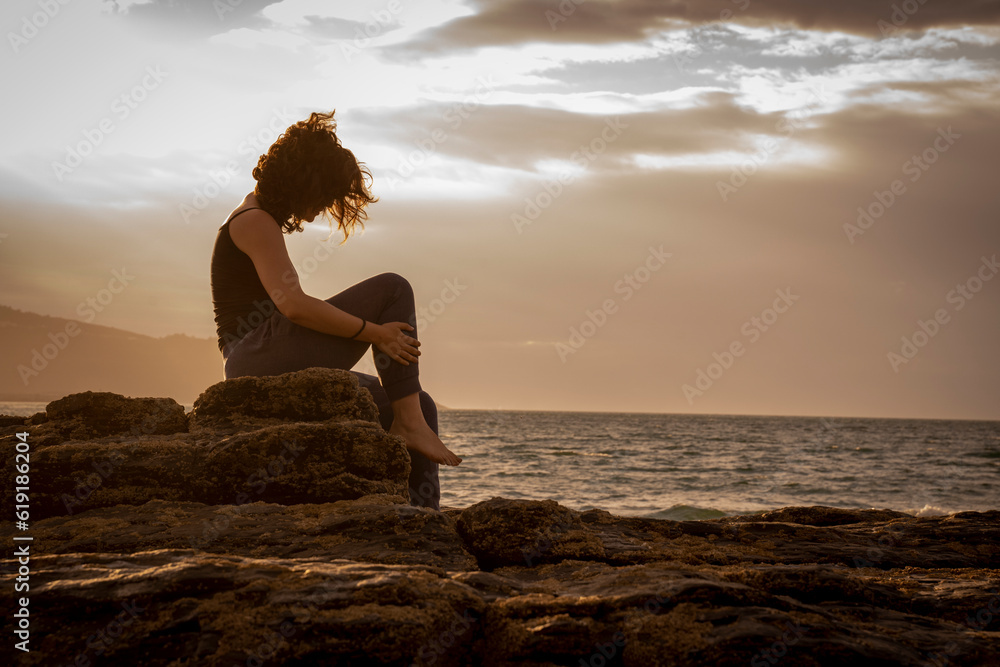 young girl sitting down comfortly on the rocks in the beach at sunset ...