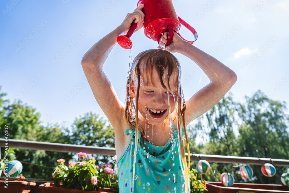 Smiling girl pouring water on head from red watering can in balcony ...