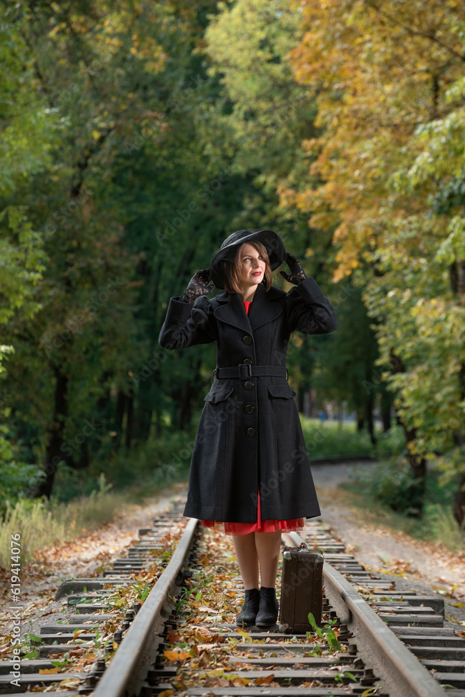 Woman in retro clothes black coat and broad-brim hat stands with old valise on rails. Woman ...