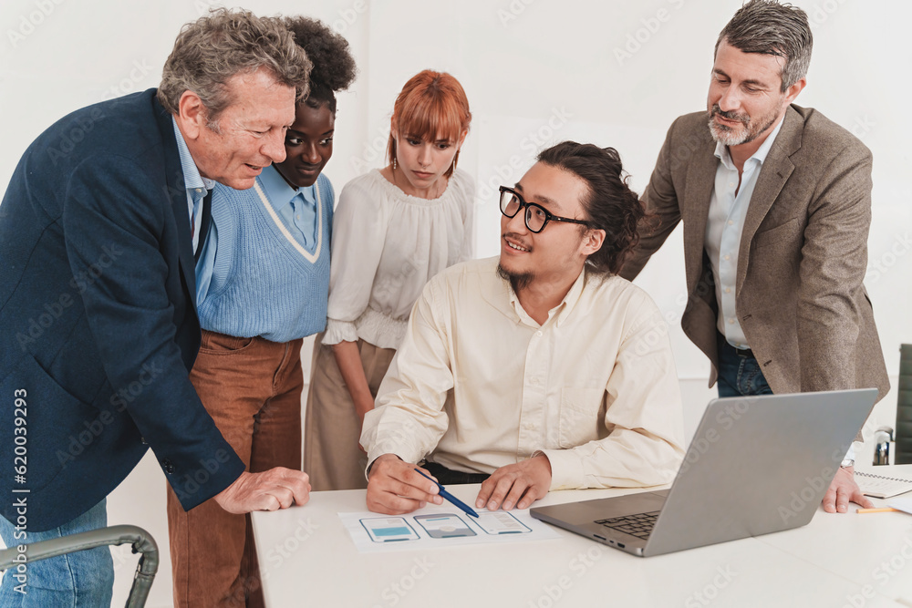 A multicultural team gathers around a young Chinese man's desk as he ...