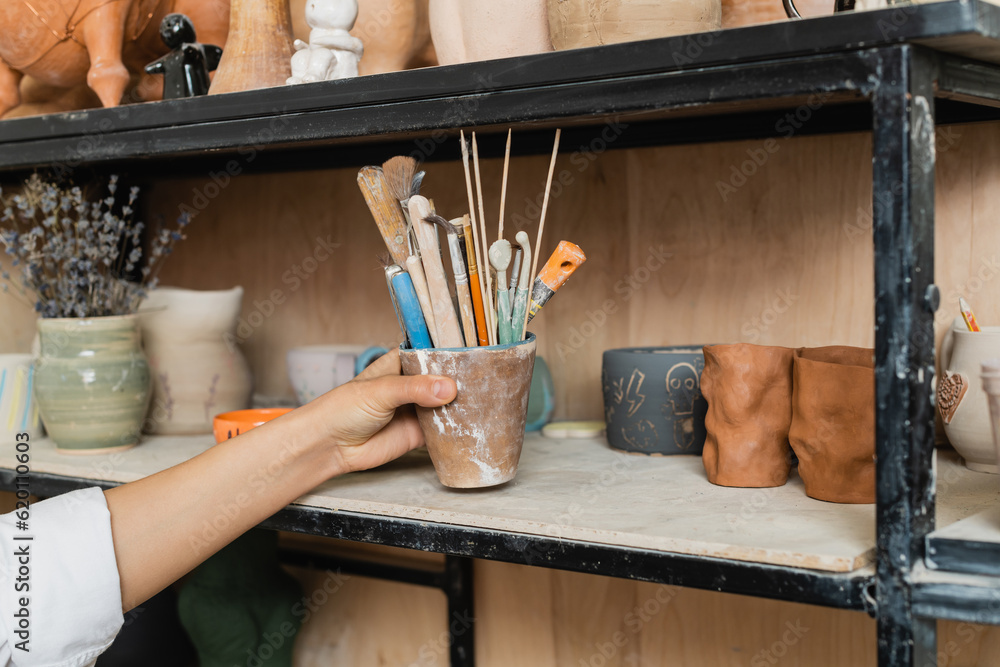 Cropped view of young craftswoman taking pottery tools from shelf with ...