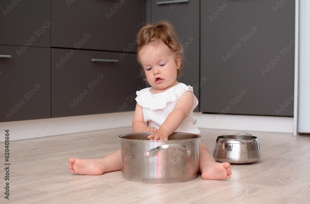 Funny little girl is sitting on kitchen floor and playing with pots ...