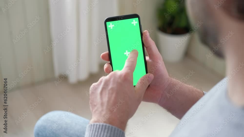 Young man sitting at home holding smartphone green mock-up screen in hand. Male person using ...