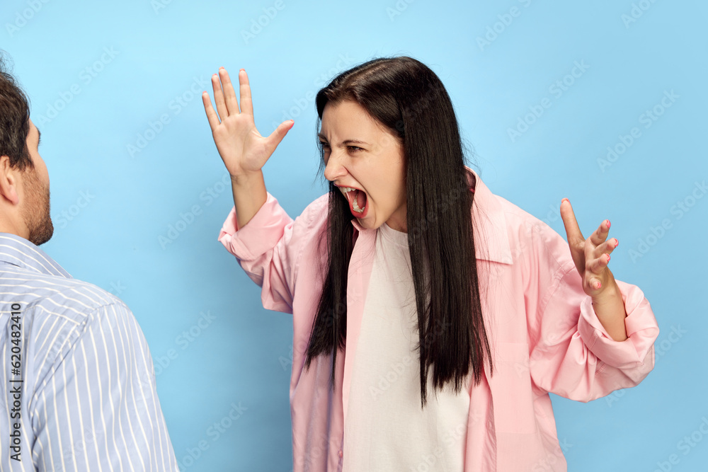 Emotional young woman loudly shouting, arguing with man against blue studio background ...