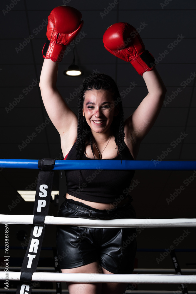 Smiling victorious female boxer in a boxing ring with her arms raised ...