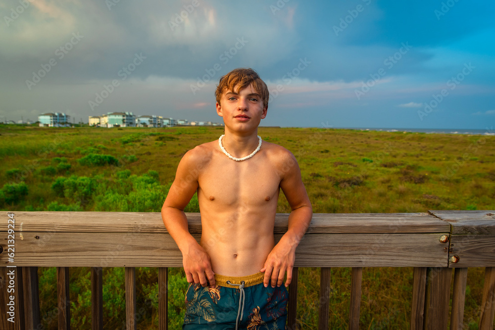 Fit teenage beach boy in swim trunks standing on a bridge overlooking ...