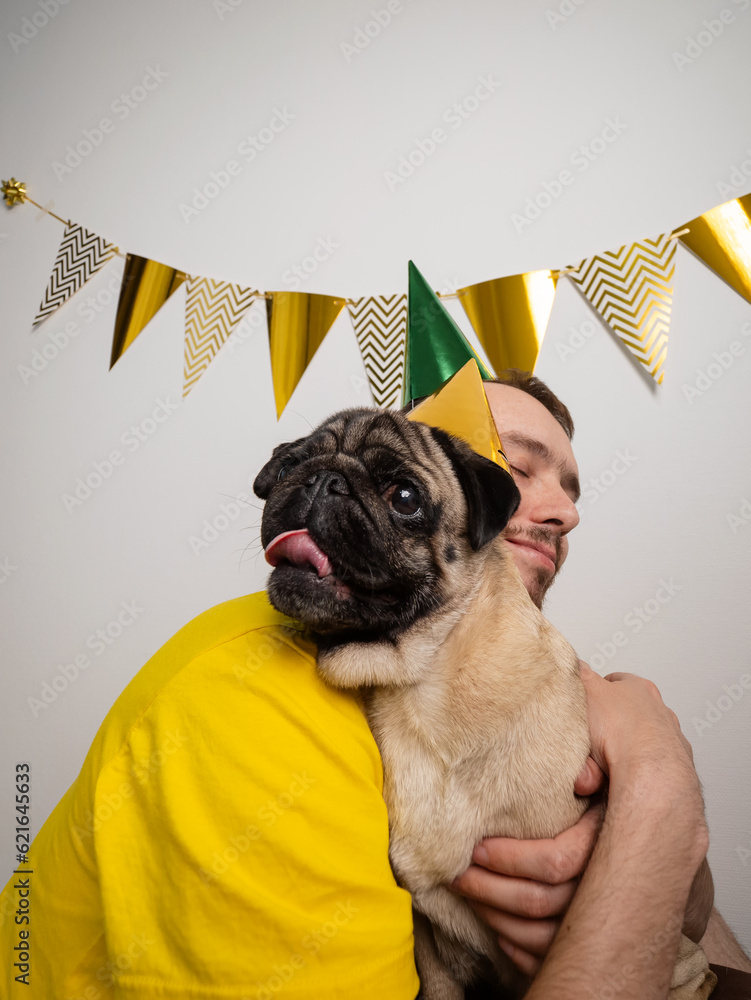 Funny guy with cute pug dog in a party hat hugging and celebrating pet ...