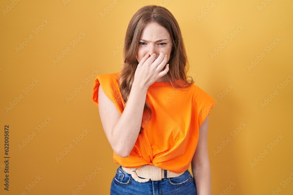 Caucasian woman standing over yellow background smelling something ...