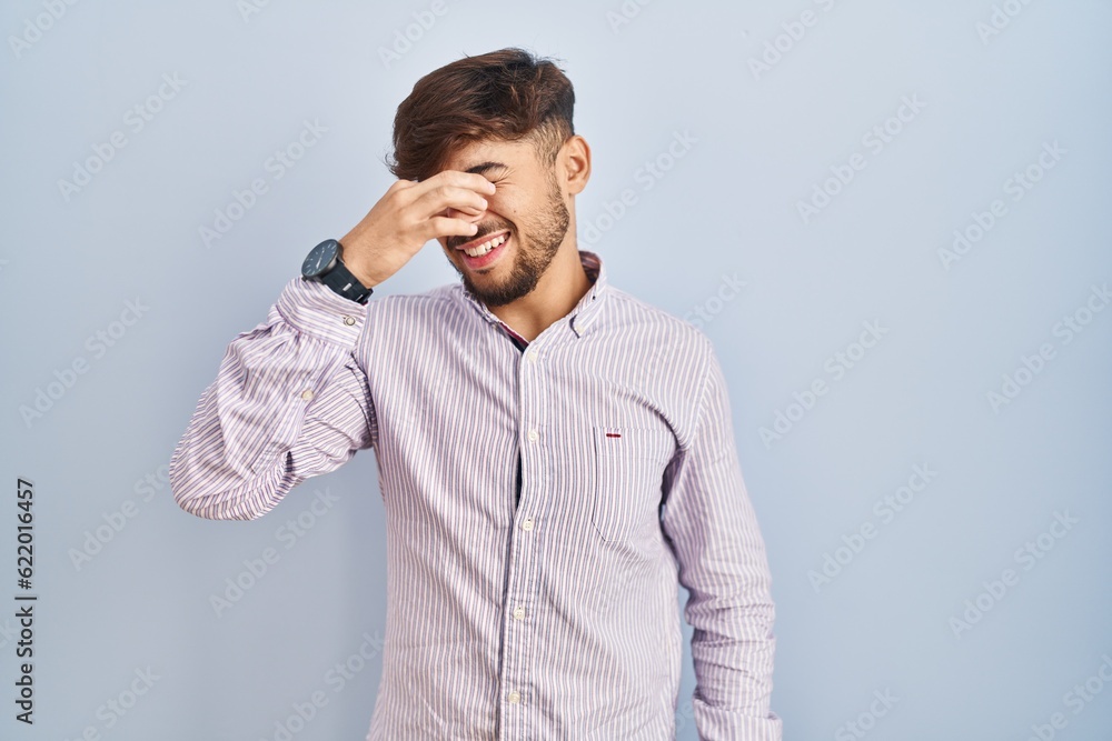 Arab man with beard standing over blue background smelling something ...