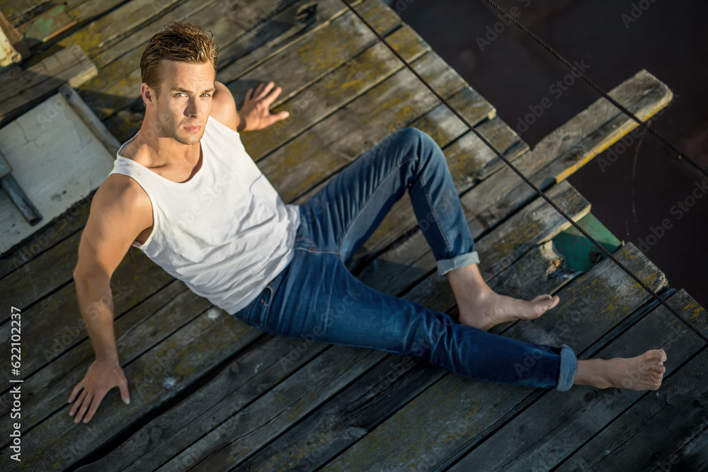 Cute barefoot guy lies on the wooden surface outdoors. He wears a white ...