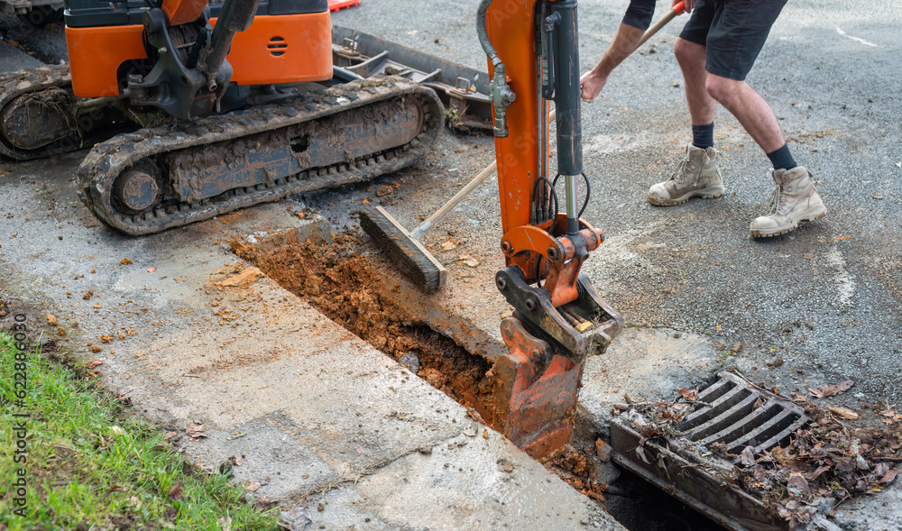 Excavator digging up dirt between cut concrete pavement. Man sweeping ...