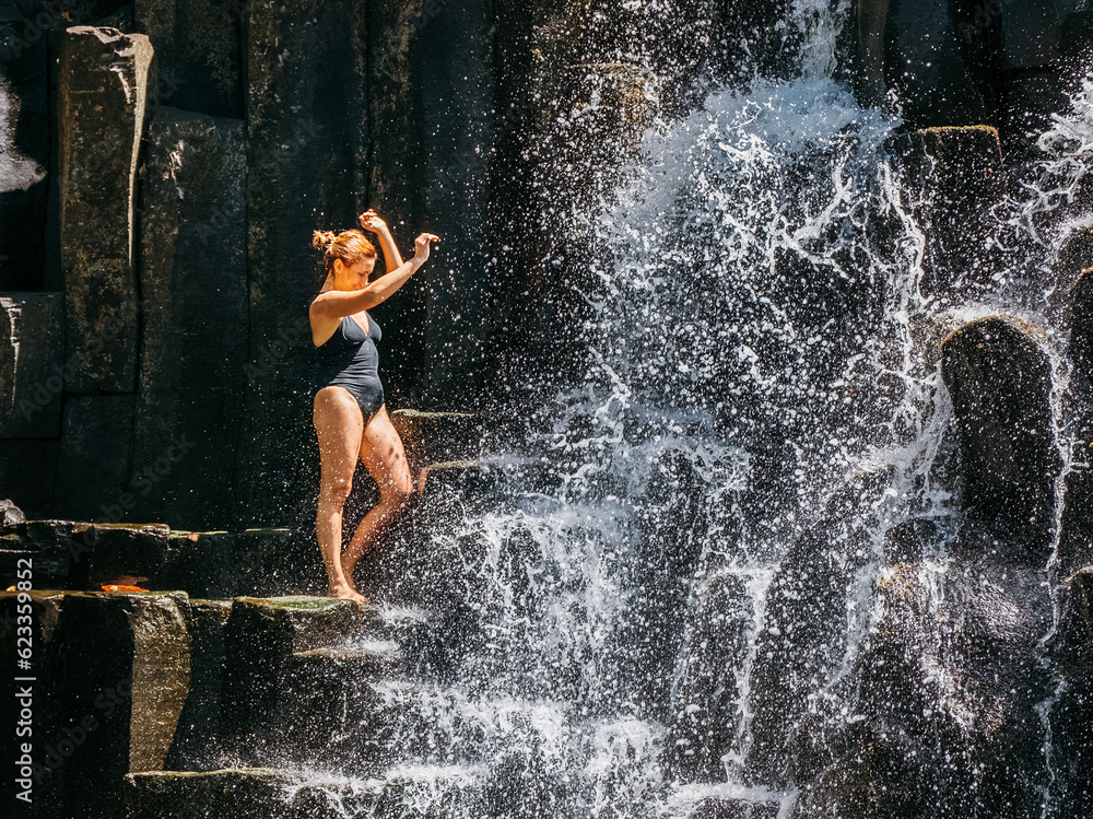 Caucasian woman in a black swimsuit refreshing under falling water streams flowing on black ...