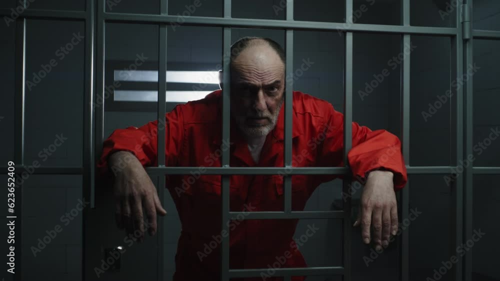Elderly prisoner in orange uniform holds hands on metal bars, looks at ... Elderly prisoner in orange uniform holds hands on metal bars, looks at ...