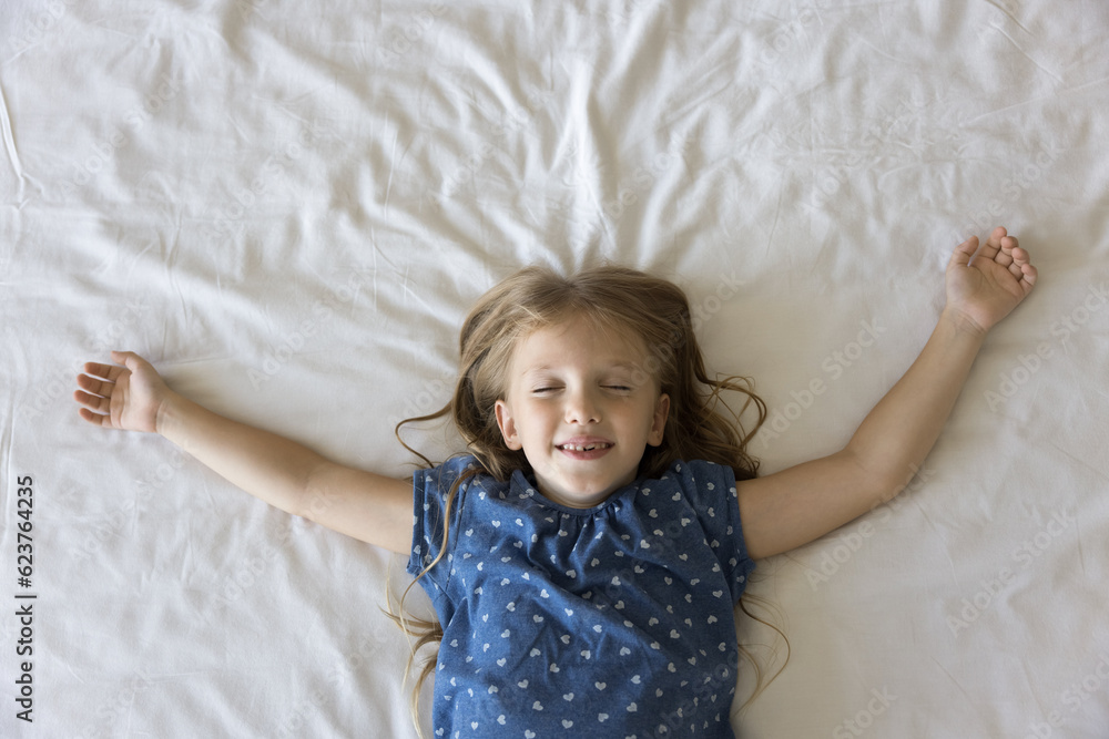 Happy sleepy preschool child girl lying on white sheet on bed with closed eyes, open arms ...