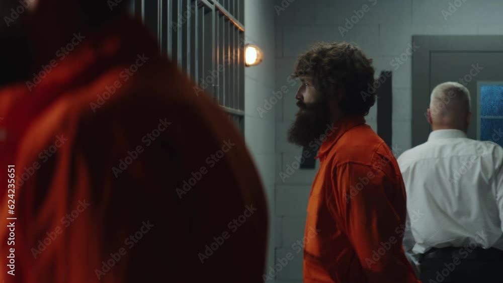 Two prisoners in orange uniforms stand facing the metal bars in front ...