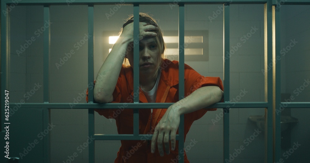 Female prisoner in orange uniform holds hands on metal bars, walks ...