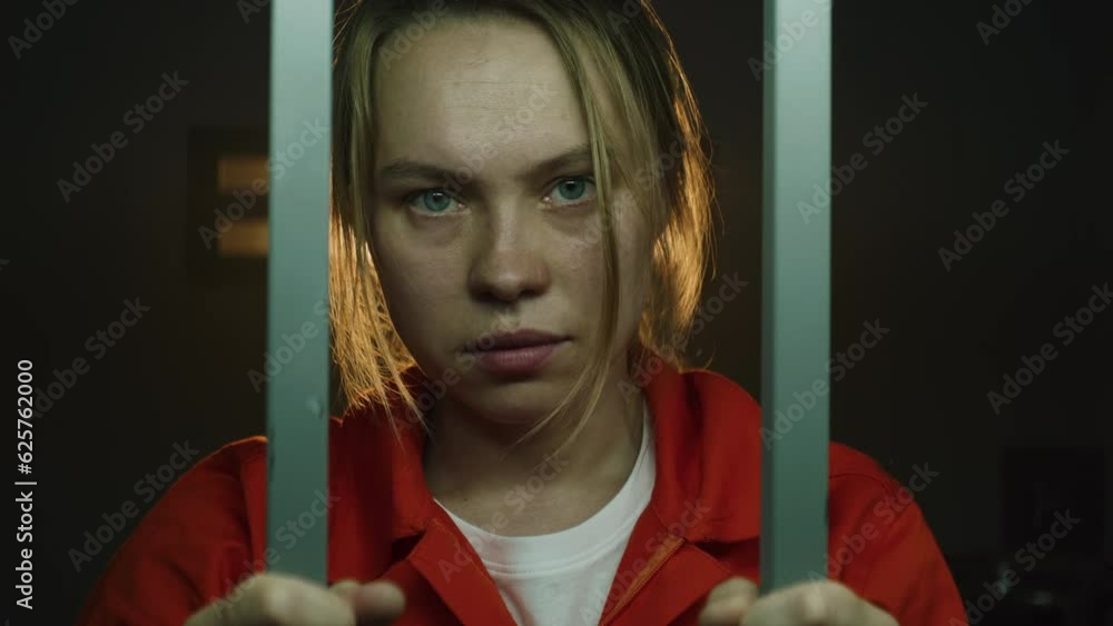 Female prisoner in orange uniform shakes, holds metal bars, looks at ...