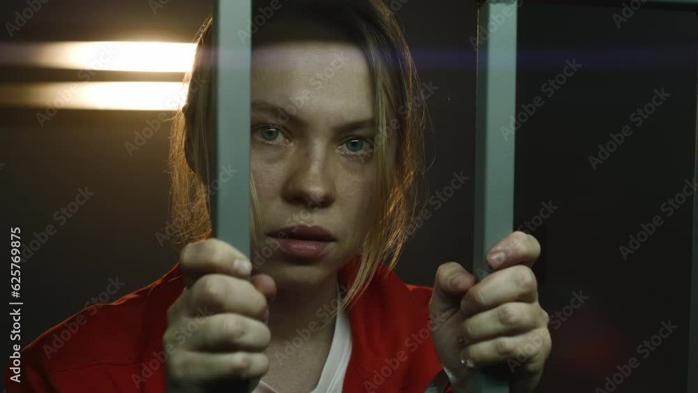Scared female prisoner in orange uniform holds metal bars, stands in ...