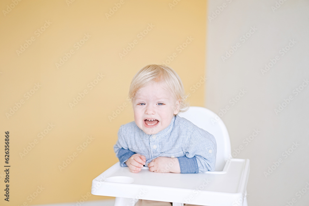 Sad upset baby is sitting in a white high chair for feeding. Little boy ...