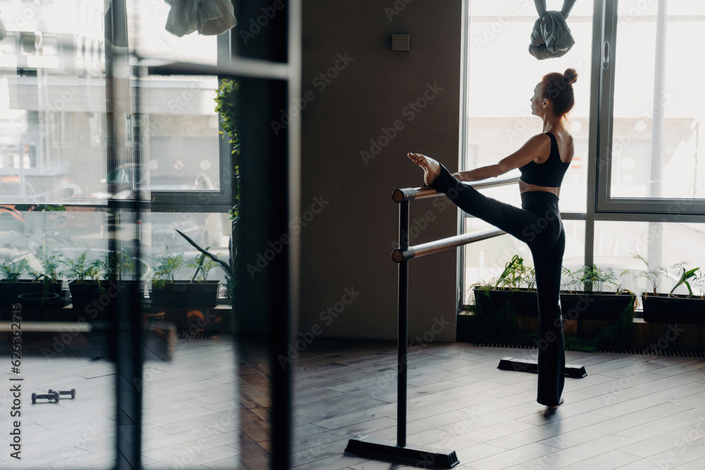 Slim red-haired ballerina stands in split position beside ballet barre ...