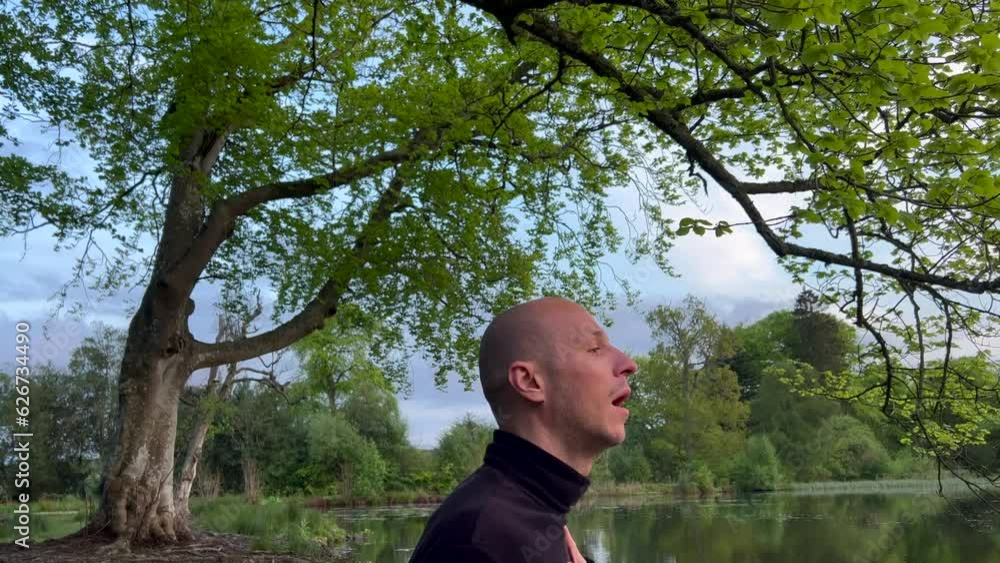 Young bald man breathwork near lake. Hand on heart under tree. Wim Hof ...