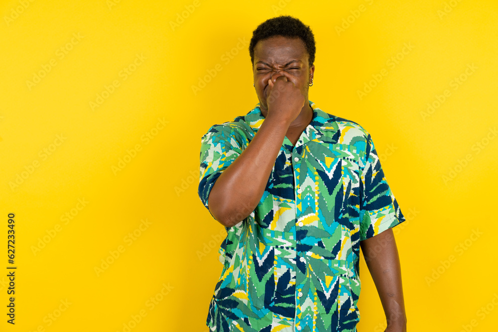 Young latin man wearing hawaiian shirt over yellow background smelling ...