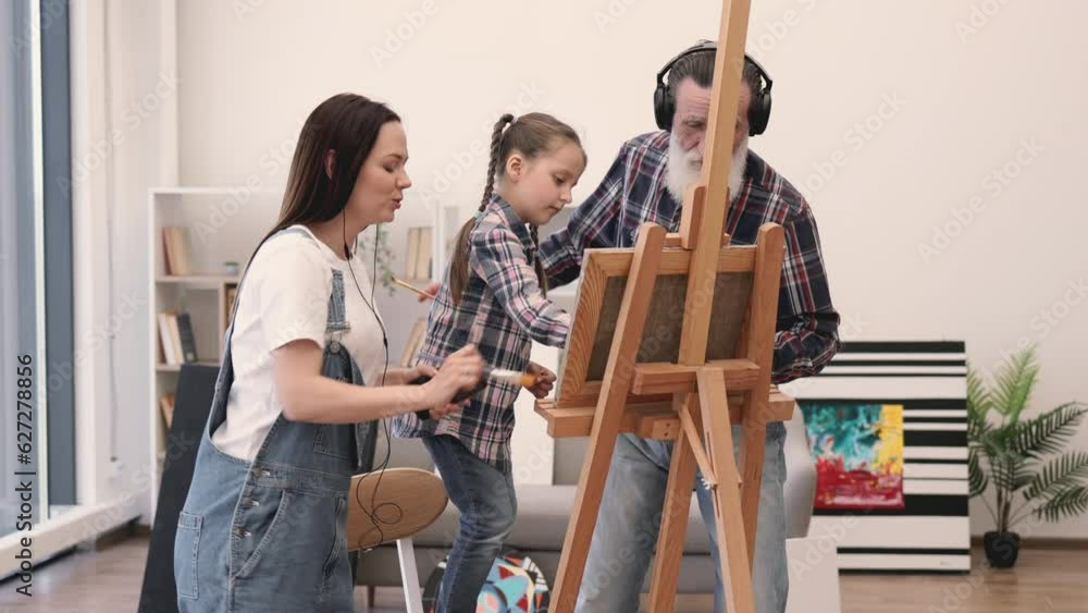 Relaxed woman with earbuds making collage on canvas with happy daughter ...