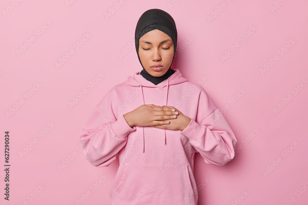 Horizontal shot of calm Muslim woman presses hands to chest has eyes ...