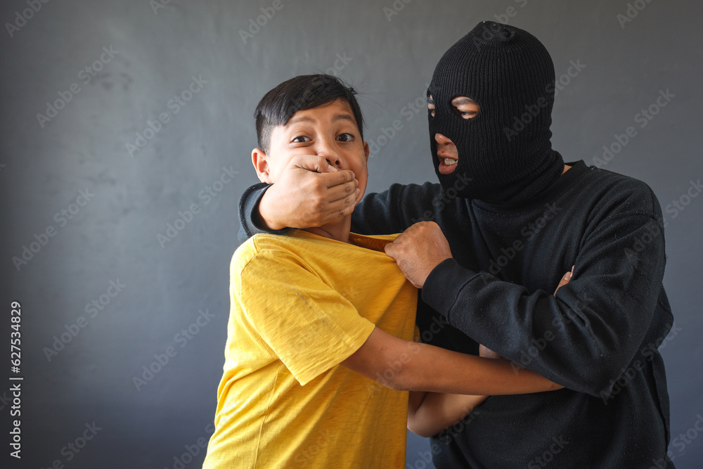 Terrible criminal man wearing black clothes and robber mask holds a little boy hostage, covering ...