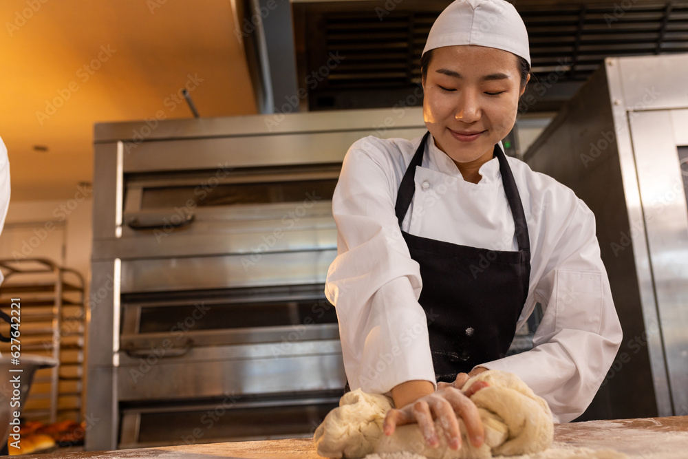 Happy asian female baker wearing apron in bakery kitchen and kneading ...