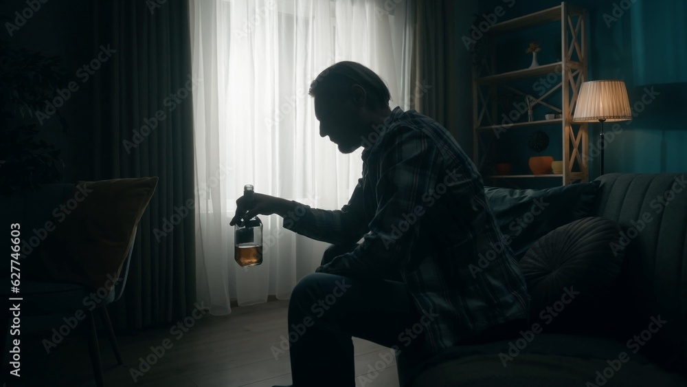 A devastated man sits in a dark room with a bottle of alcohol. Side ...