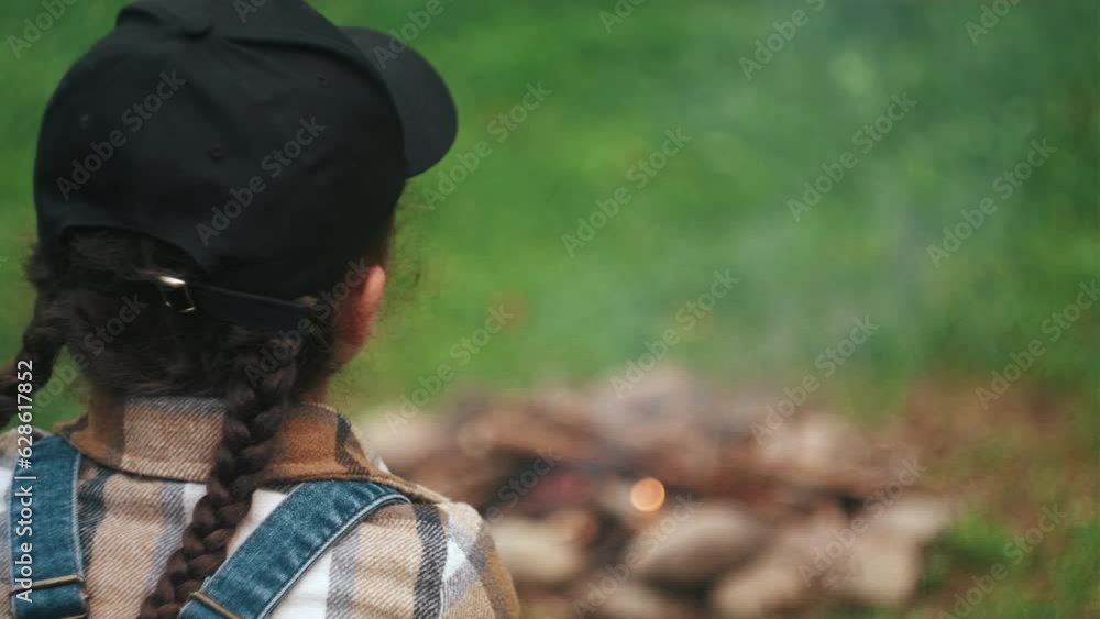 Selective focus of preteen girl in cap sitting near bonfire and look at fire. Little child lit ...