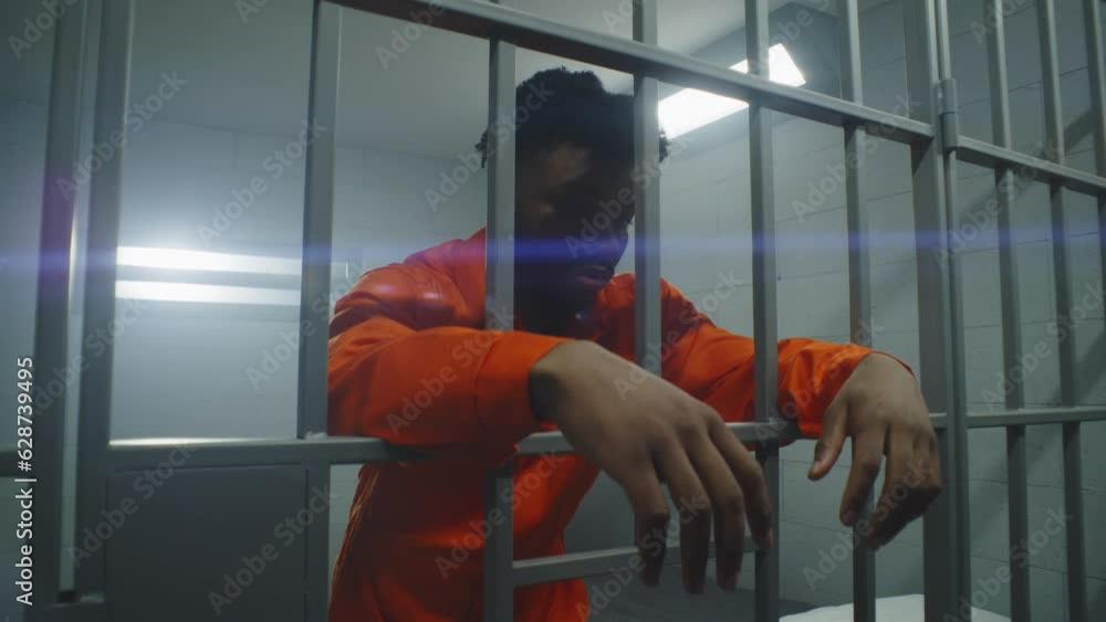 African American prisoner in orange uniform leans on jail cell bars and ...