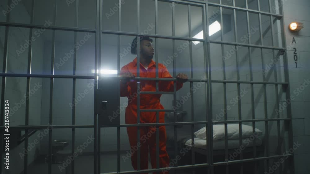 African American prisoner in orange uniform leans on jail cell bars and ...