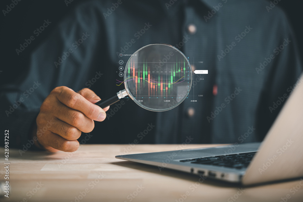 Hand of a businessman or trader holds a magnifier glass, examining the stock market chart for ...