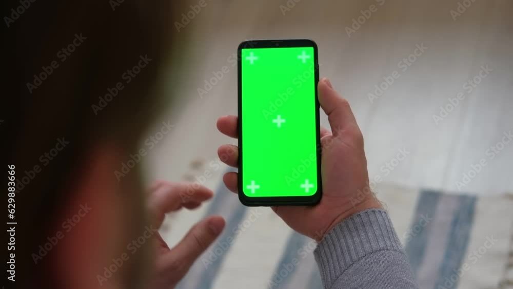 Young man sitting at home holding smartphone green mock-up screen in hand. Male person using ...