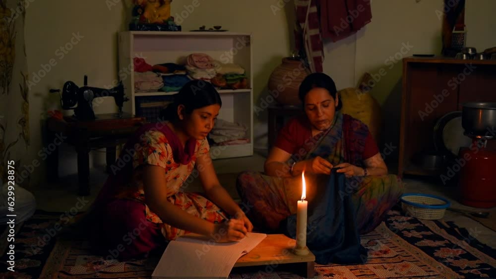 A teenage girl is completing her homework while her mother is stitching ...
